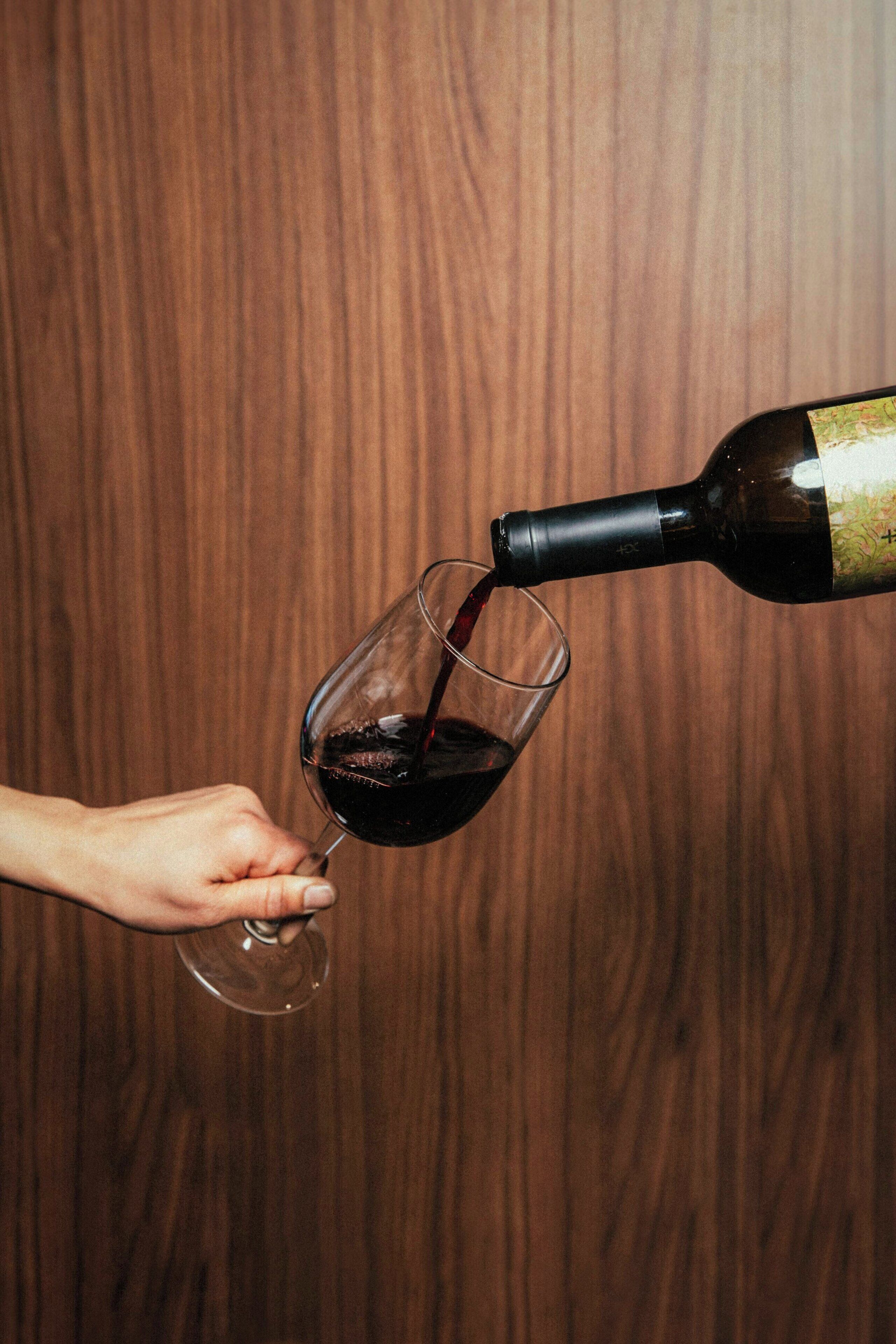 Uma mão segurando uma taça de vinho recebendo vinho tinto de uma garrafa.