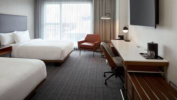 Quarto de hotel com duas camas, uma mesa, uma cadeira, televisão e uma janela grande com cortinas.