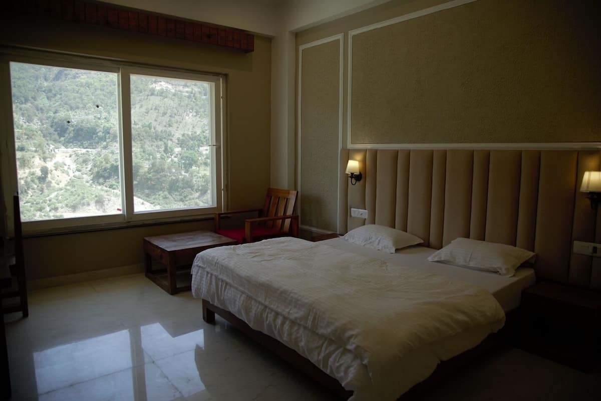 Quarto com cama grande, uma cadeira, uma mesinha e janela com vista para área verde.