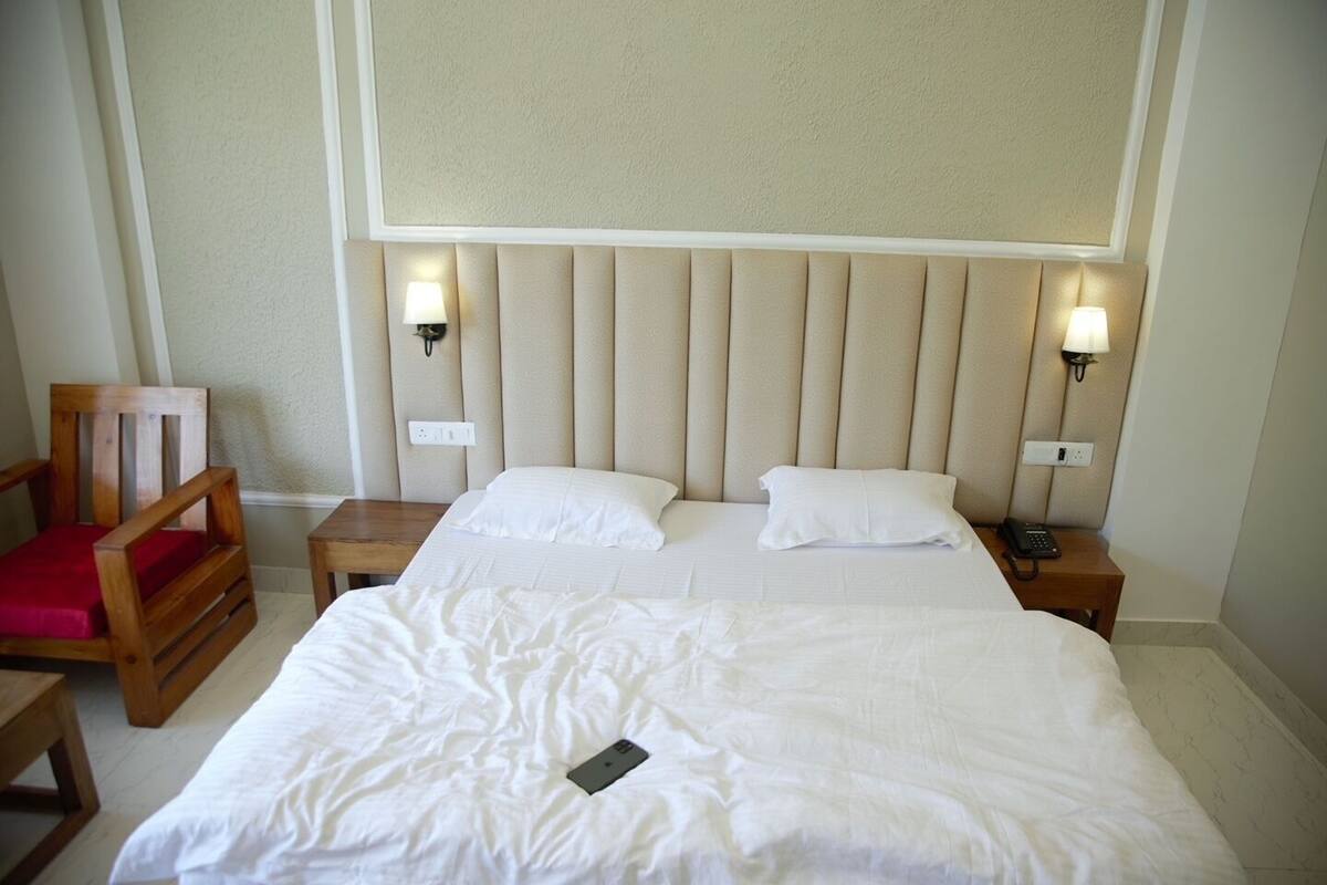 Quarto de hotel com uma cama, duas luminárias de cabeceira, uma cadeira de madeira e uma mesinha de cabeceira.