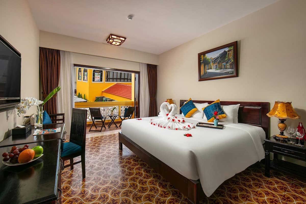 Quarto de hotel com uma cama grande, uma escrivaninha com cadeira, um espaço de refeições com mesa e cadeiras, e vista para a rua através de uma janela.