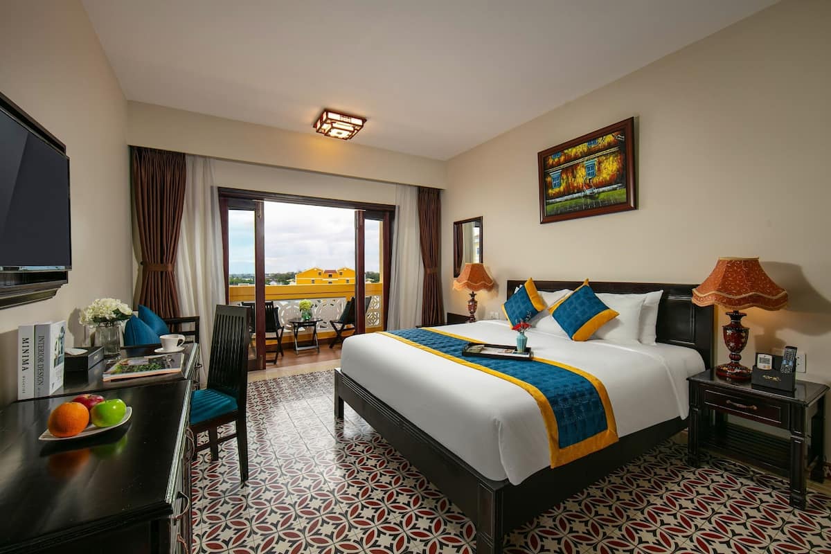 Quarto de hotel com uma cama grande, uma secretária com computador, uma cadeira, uma televisão e uma janela com vista para edifícios.