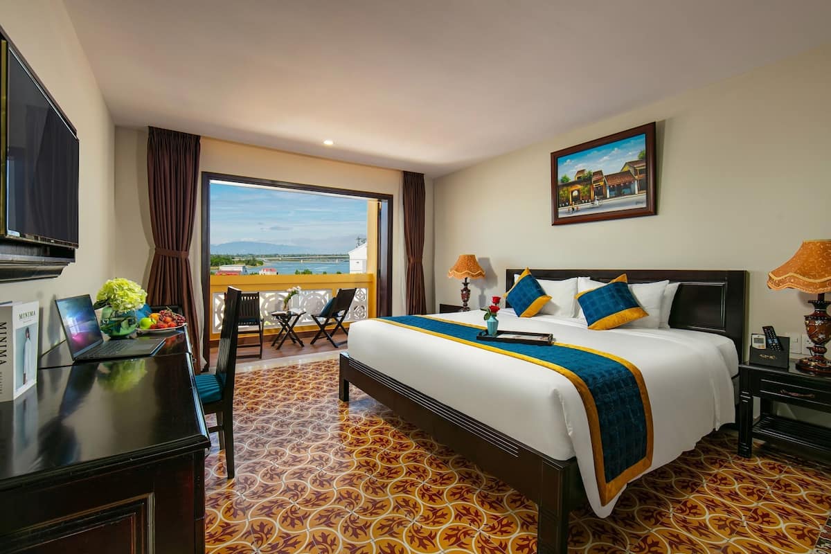 Quarto de hotel com uma cama grande, uma escrivaninha com um laptop, uma cadeira, varanda com vista e um quadro na parede.