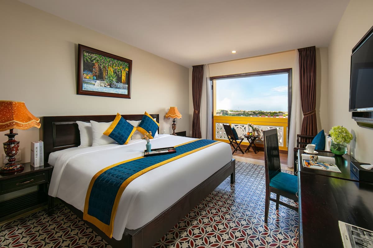 Quarto de hotel com uma cama grande, uma escrivaninha com cadeira, televisão e varanda com vista.
