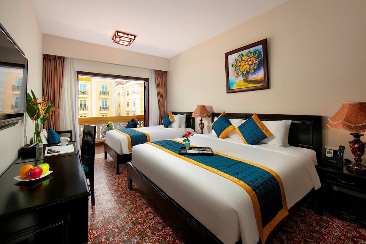 Quarto de hotel com duas camas, uma escrivaninha com frutas, uma janela com cortinas, um quadro e um tapete estampado.