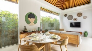 Uma sala de jantar moderna com uma mesa redonda posta para quatro pessoas, cadeiras de madeira, uma janela grande com vista para bambu e uma televisão fixada na parede.