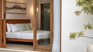 Quarto com estrutura de cama de madeira, roupa de cama branca, almofadas estampadas, vista através de uma porta de vidro e uma planta de bambu à direita.