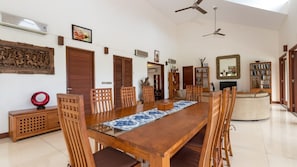 Uma sala de jantar espaçosa com uma longa mesa de madeira, cadeiras e um corredor azul estampado. Há ventiladores de teto, uma estante e uma lareira ao fundo.