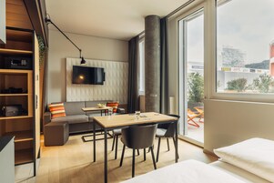 Quarto de hotel moderno com sofá, mesa de jantar, cadeiras, TV fixada na parede e uma grande janela com vista para a cidade.