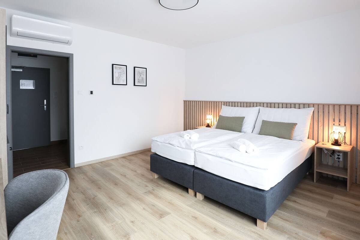 Quarto de hotel moderno com cama de casal, mesas de cabeceira com luminárias, uma poltrona cinza e piso de madeira.