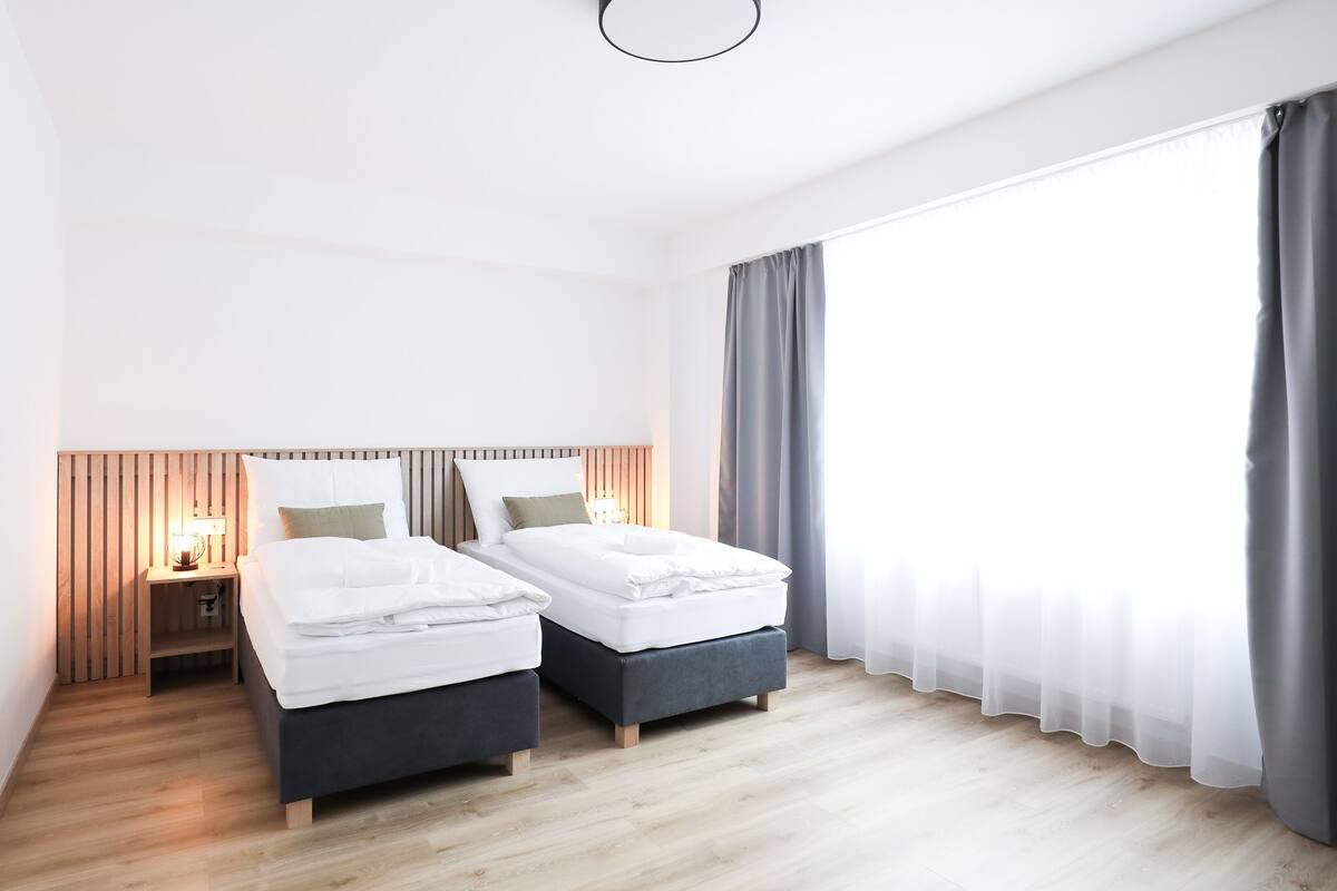 Quarto moderno com duas camas, cabeceira de madeira, mesa de cabeceira com abajur e uma janela grande com cortinas.