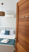 Quarto de hotel com porta de madeira, cama com roupa de cama branca e travesseiro azul, radiador branco e luminária pendente.