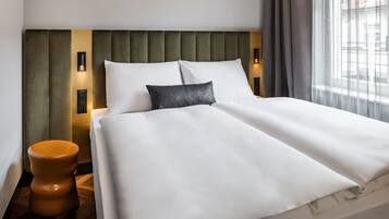 Uma cama bem arrumada com lençóis brancos, uma cabeceira estofada em verde e um banquinho laranja ao lado.