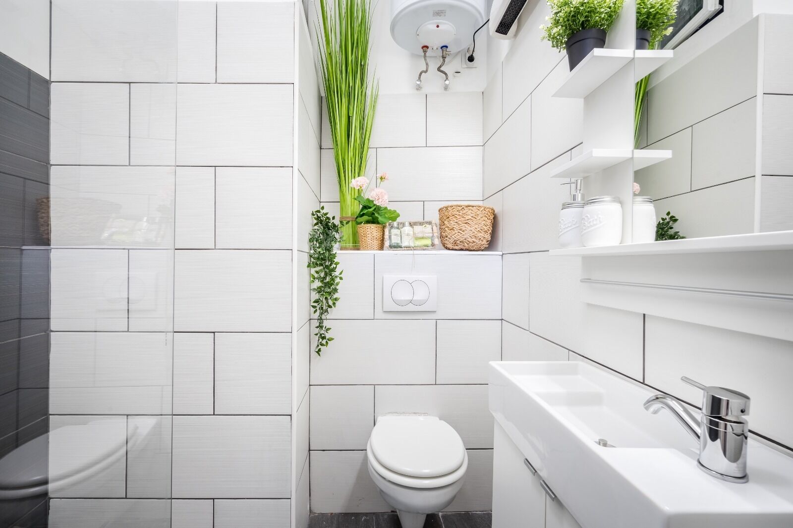 Casa de banho moderna com azulejos brancos, sanita, lavatório e estantes com plantas e artigos de higiene pessoal.