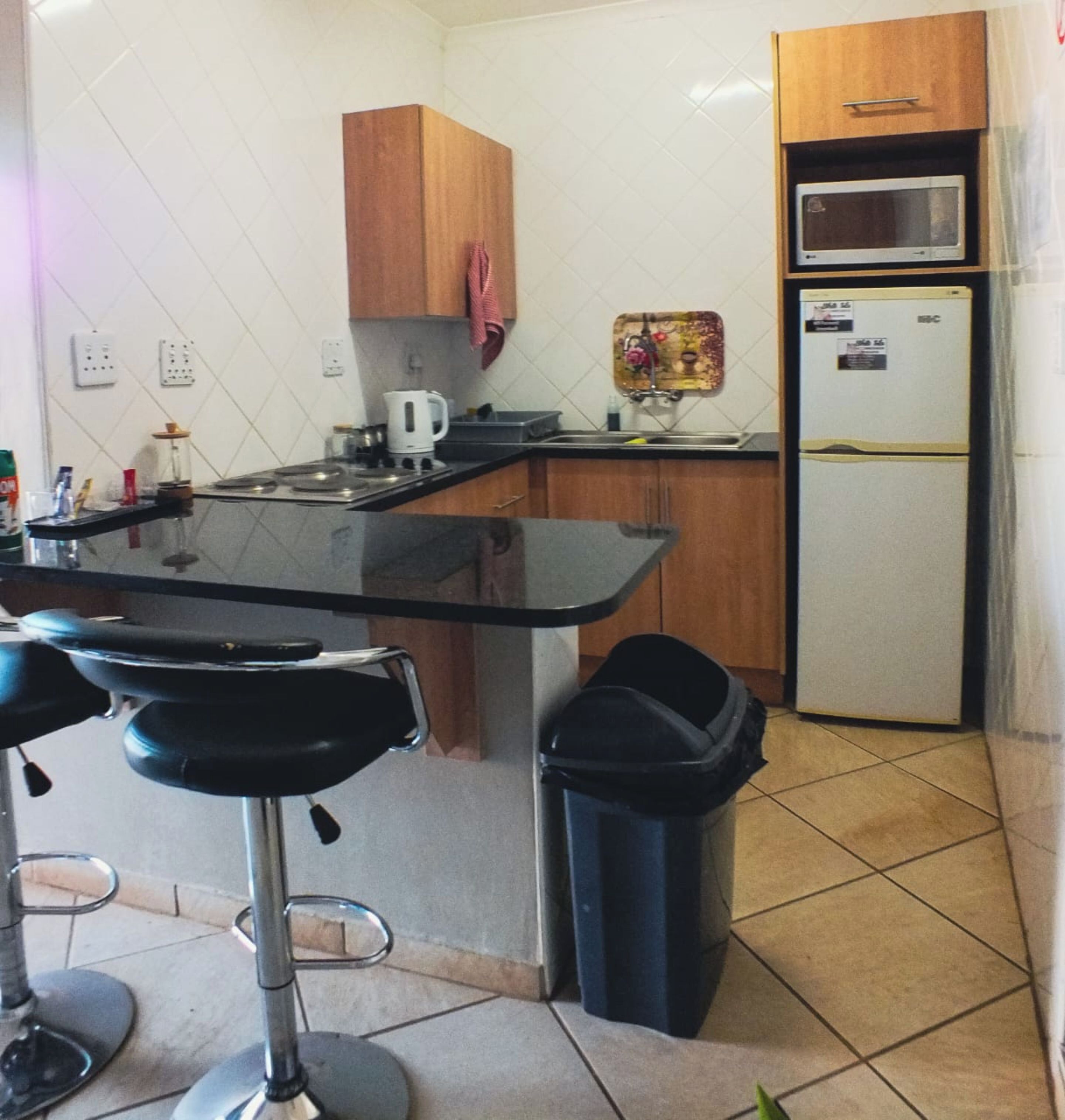 Uma cozinha com bancada preta, armários de madeira, pia de aço inoxidável, micro-ondas e um cesto de lixo.