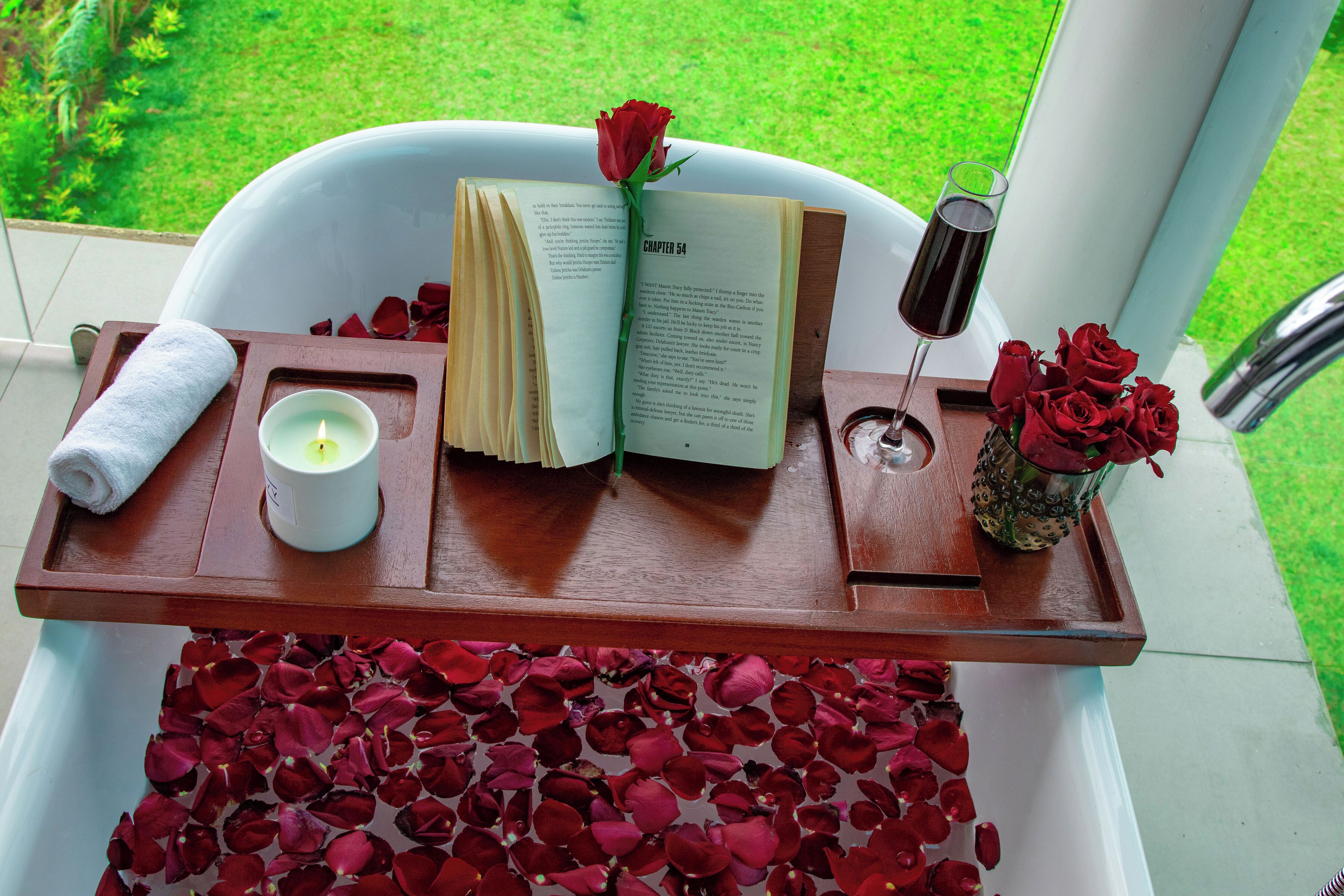 Uma banheira com pétalas de rosa, um livro, uma vela e um copo com líquido vermelho.