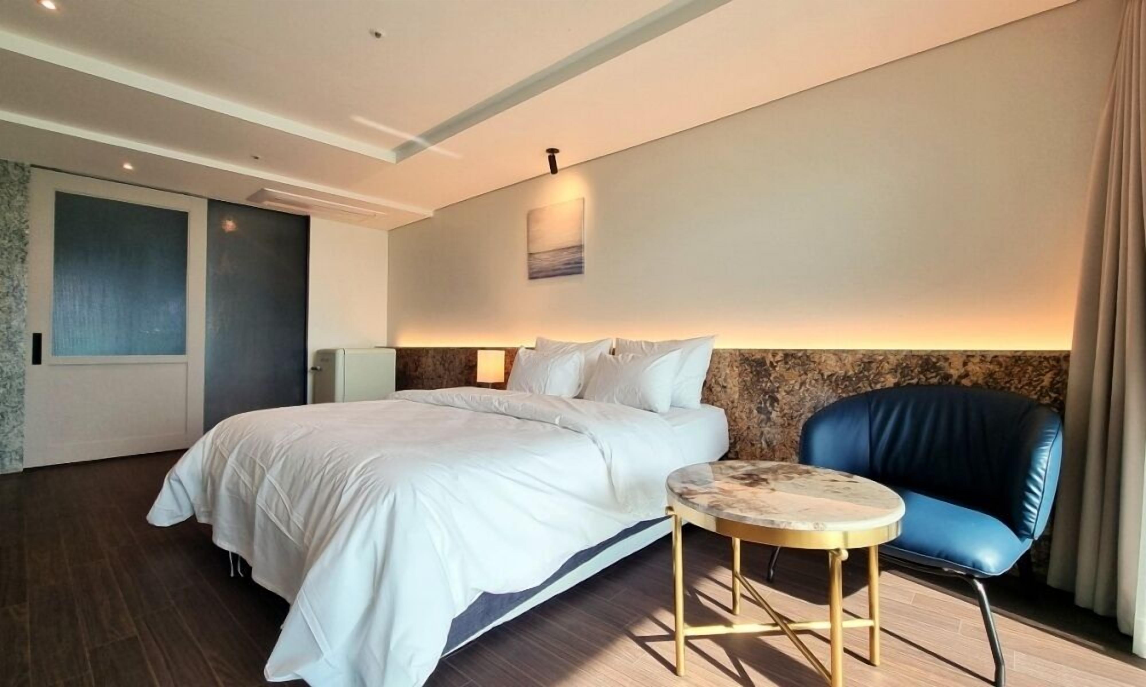 Quarto de hotel moderno com uma cama grande, uma mesa redonda pequena, uma cadeira azul e uma luminária fixada na parede.