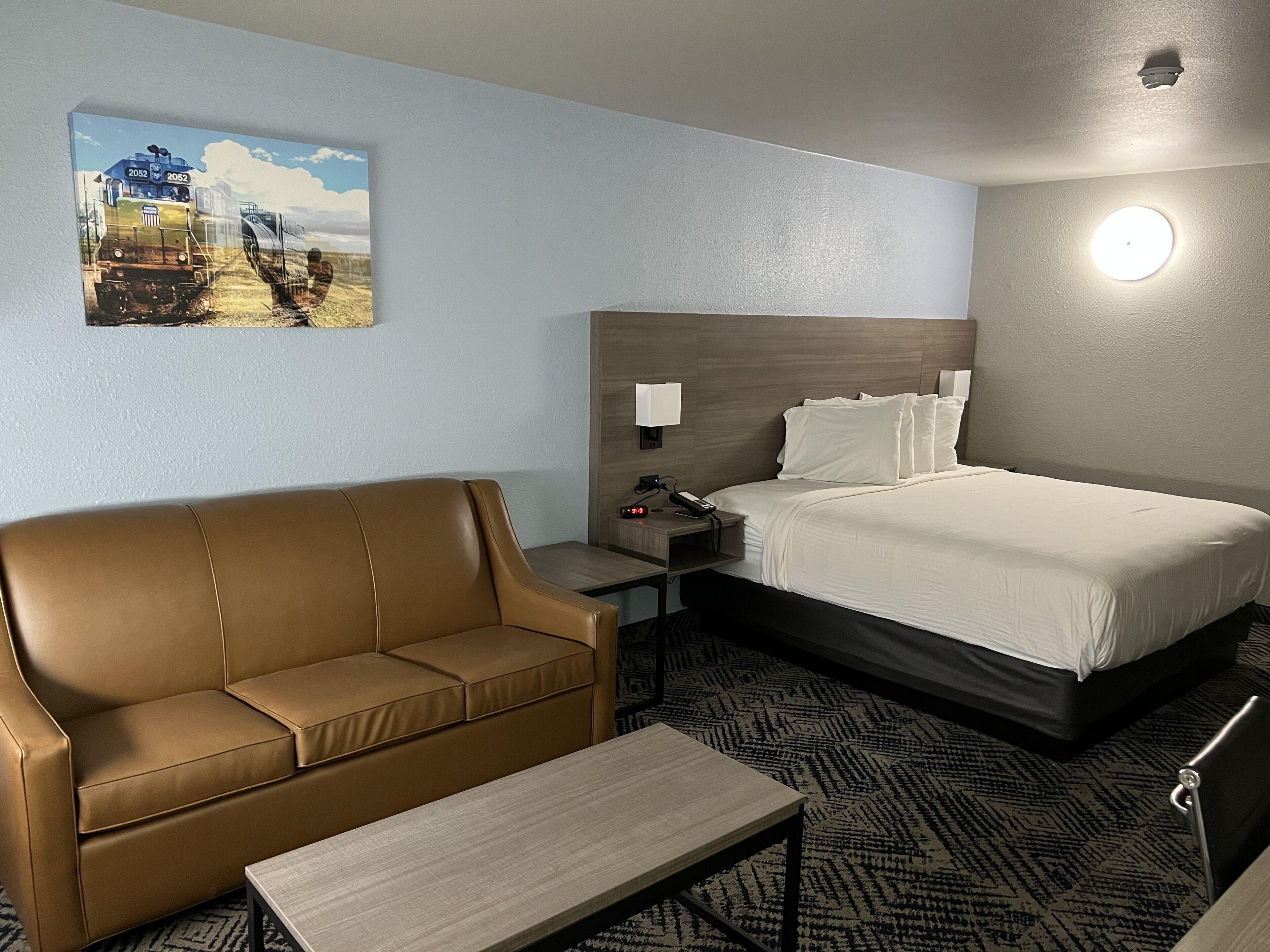 Quarto de hotel com uma cama, um sofá, uma mesinha, uma mesa de cabeceira, um abajur e um quadro de um trem.
