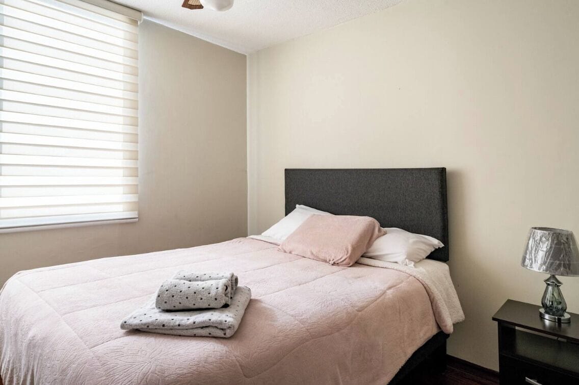 Cama bem arrumada com colcha rosa e almofadas, luminária de cabeceira e janela com persianas.