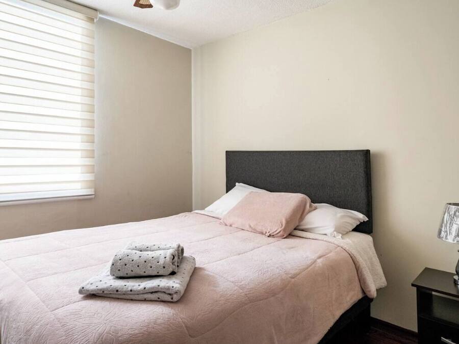 Cama bem arrumada com colcha rosa e almofadas, luminária de cabeceira e janela com persianas.