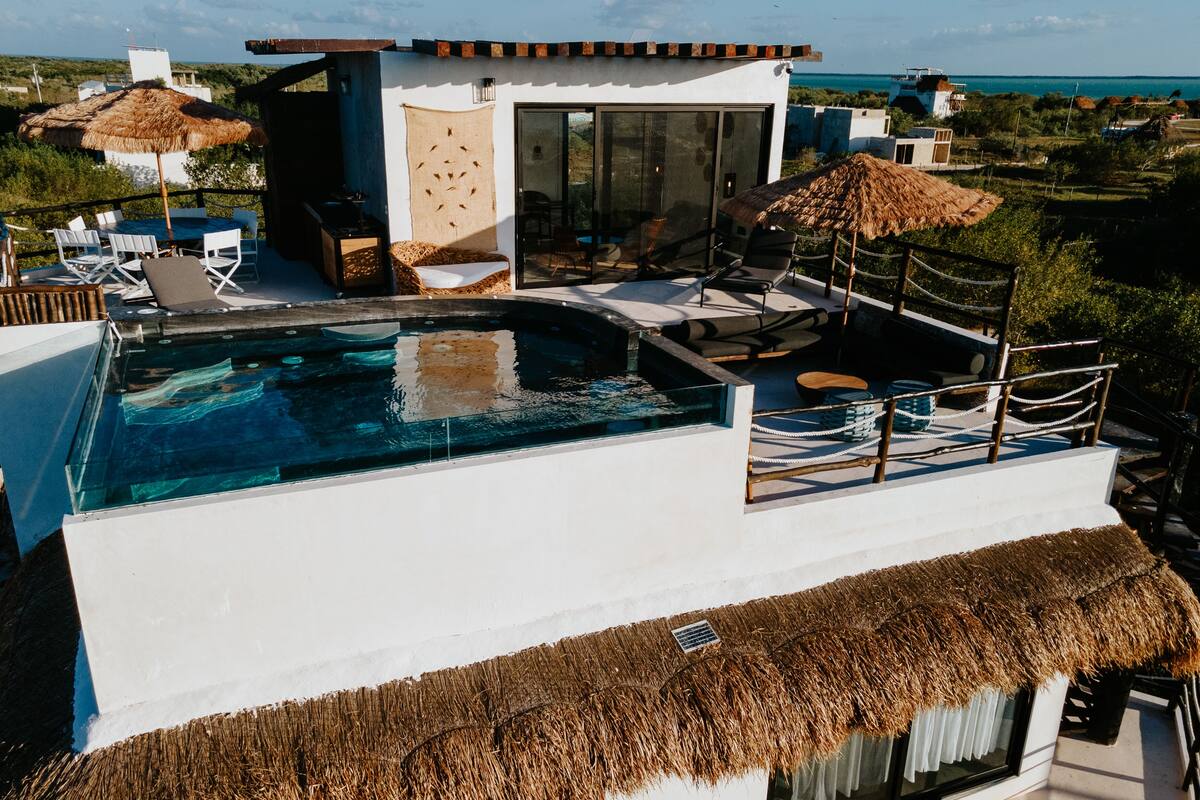 Uma piscina infinita no terraço com um telhado de palha, cadeiras de descanso e uma área de estar coberta de vidro com vista para o oceano.