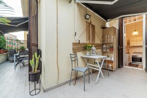 Uma área de pátio com mesa e cadeiras, um vaso de planta e uma cozinha visível através de uma porta aberta.