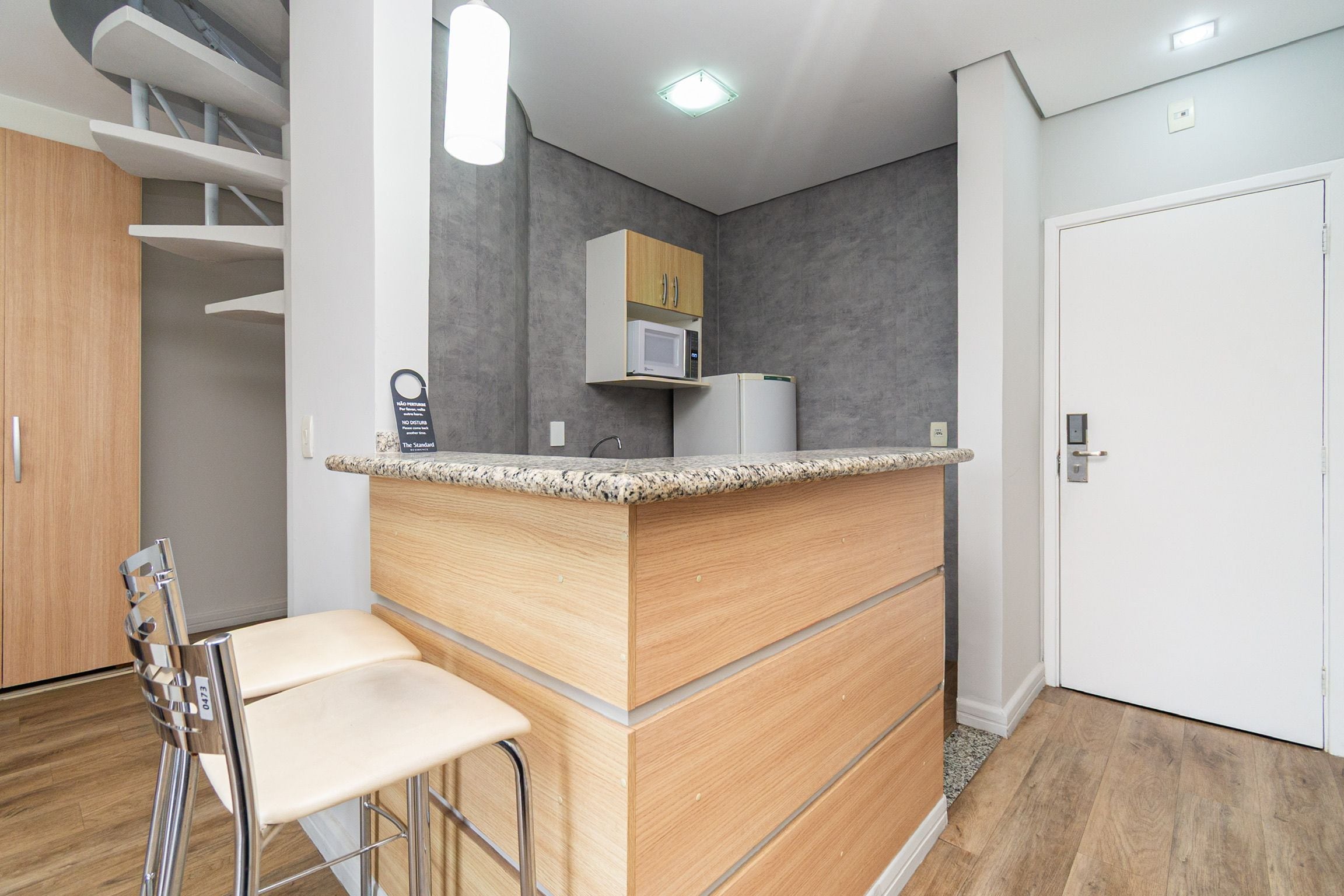 Cozinha moderna com bancada de madeira, micro-ondas e frigorífico. Há um banco de bar e uma porta à direita.
