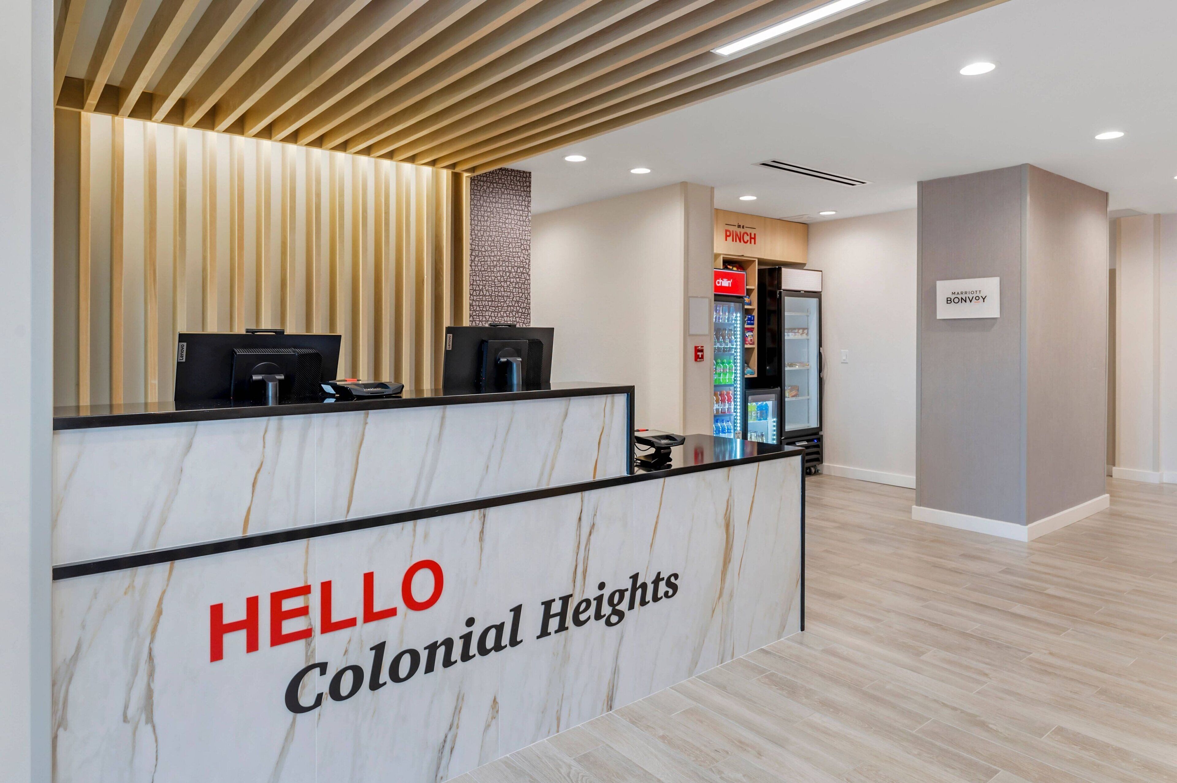 Uma área de recepção de hotel com balcão de mármore, uma placa com 'HELLO Colonial Heights', uma geladeira 'PINCH' e uma placa 'MARRIOTT BONVOY'.