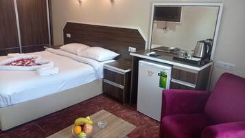 Quarto de hotel com cama, pequeno espaço de cozinha, espelho, cadeira roxa e uma fruteira em cima da mesa.