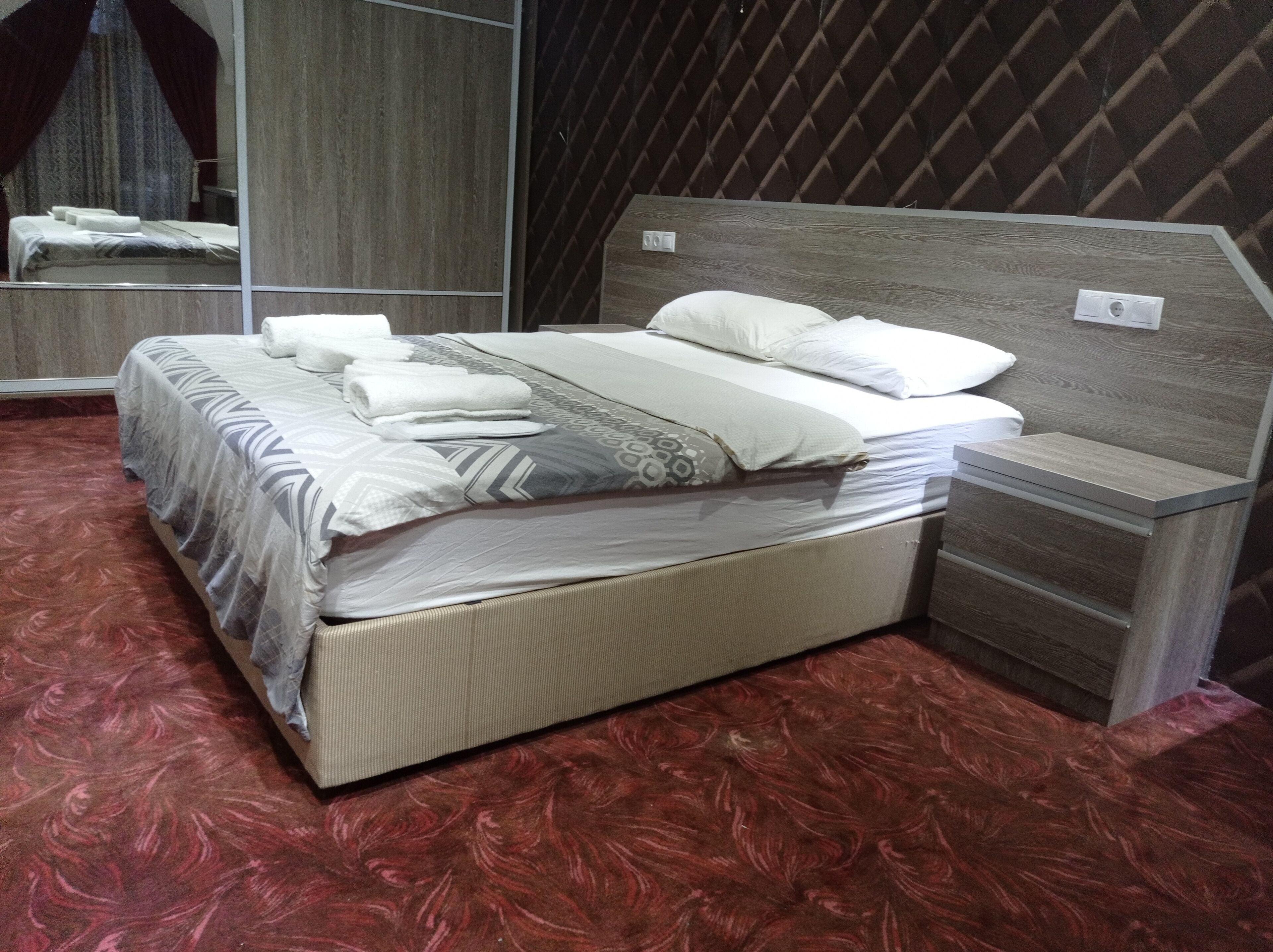 Uma cama bem arrumada com lençóis brancos e um espalha e cobertura estampada, uma mesa de cabeceira com abajur, e uma parede com padrão geométrico.
