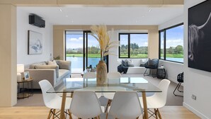 Uma sala de estar moderna com uma mesa de jantar de vidro, cadeiras brancas, um sofá e uma grande janela com vista para um lago.