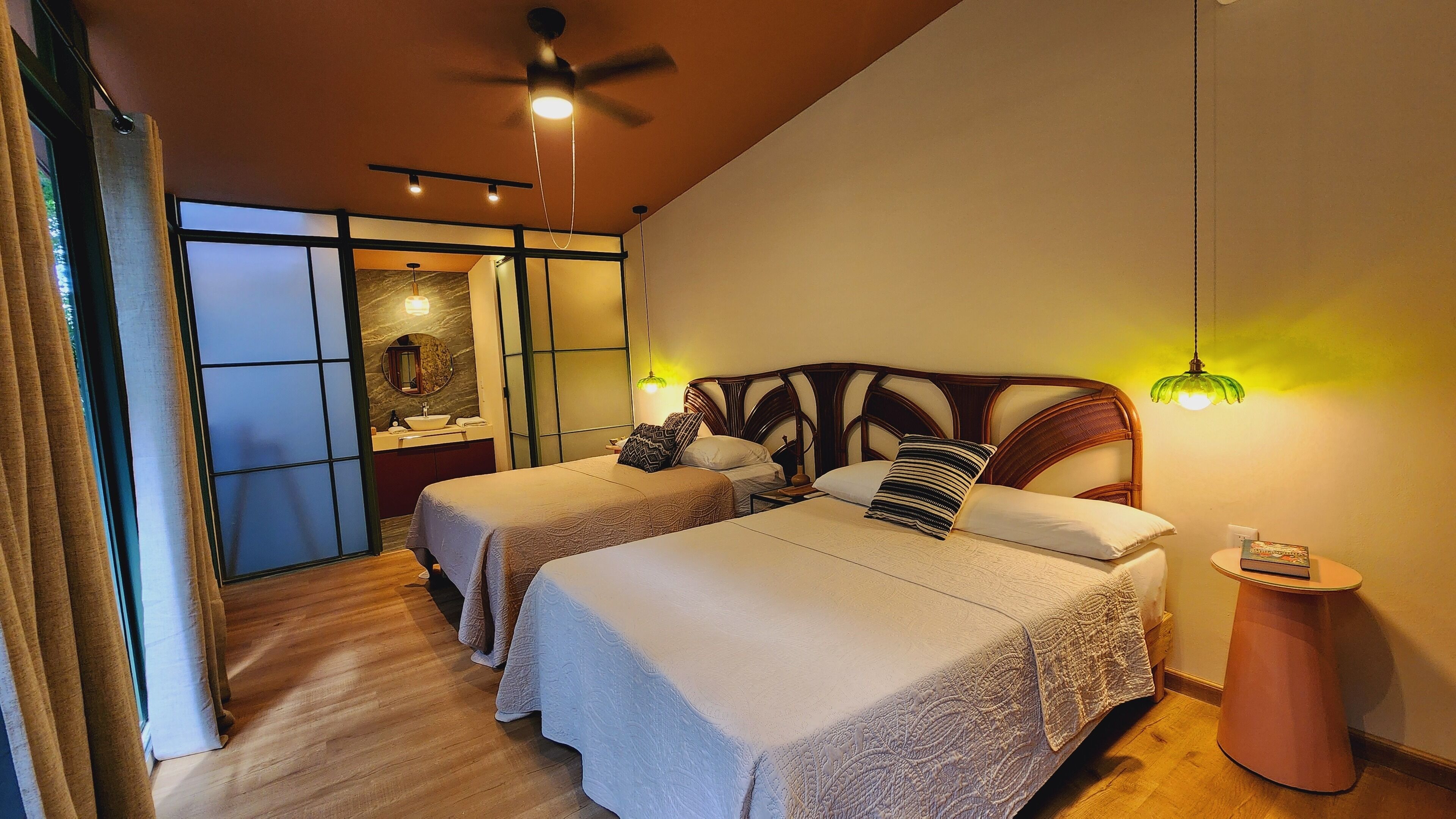 Quarto de hotel com duas camas, um ventilador de teto, uma mesa pequena com abajur e vista para o exterior por uma porta de correr.