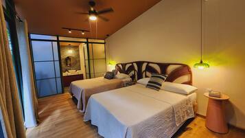 Quarto de hotel com duas camas, um ventilador de teto, uma mesa pequena com abajur e vista para o exterior por uma porta de correr.