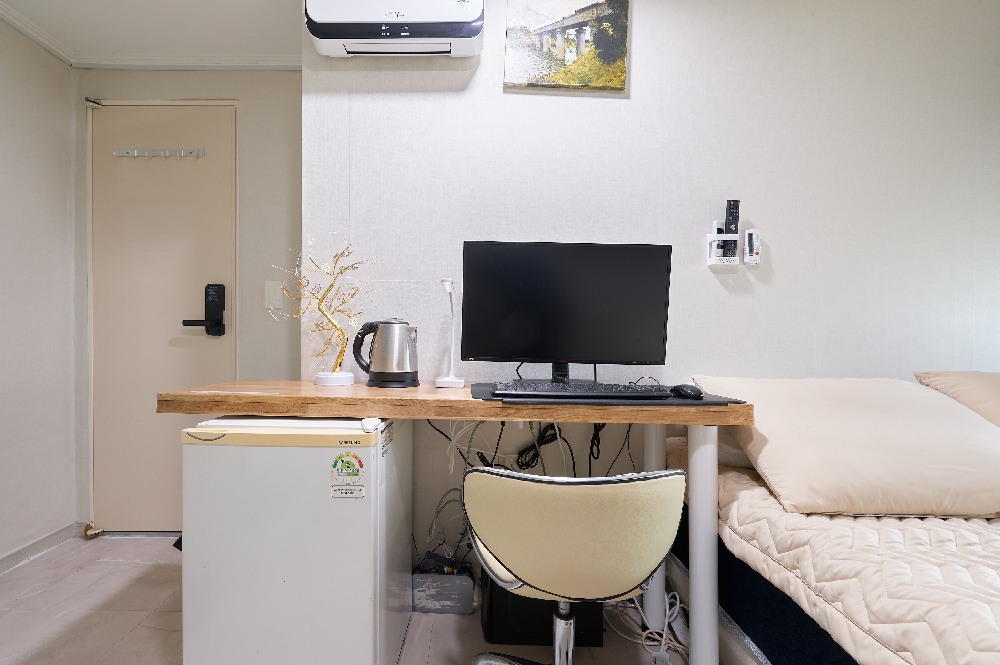 Quarto compacto com cama, escrivaninha com computador, pequeno frigorífico e uma cadeira.