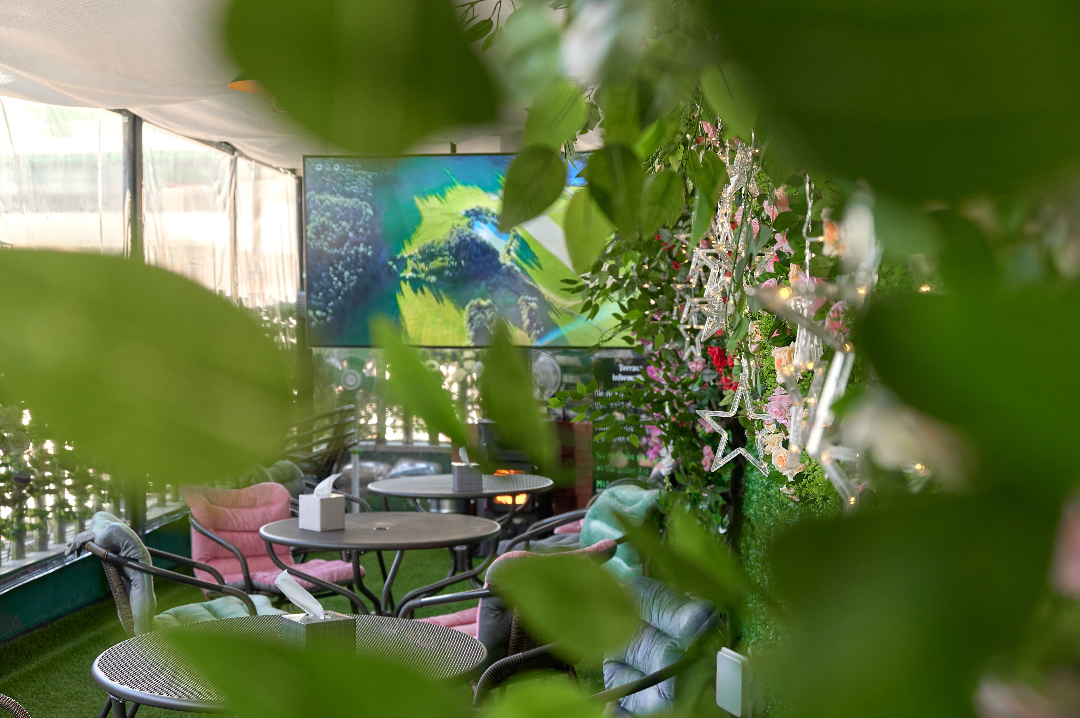 Um espaço de estar interno com mesas redondas e cadeiras estofadas, rodeado por vegetação e plantas decorativas.