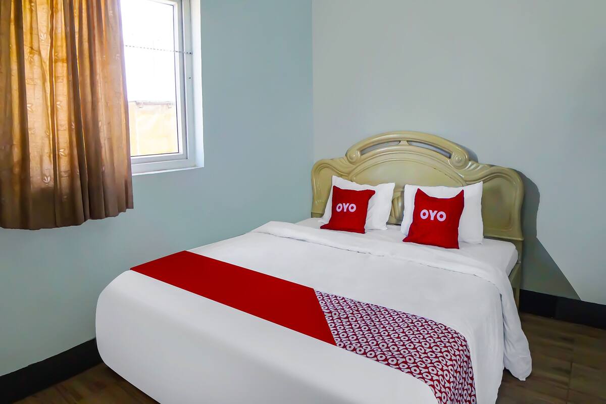 Uma cama bem arrumada com uma colcha branca e vermelha, duas almofadas com a marca 'OYO', uma janela com cortinas marrons e uma parede azul claro.