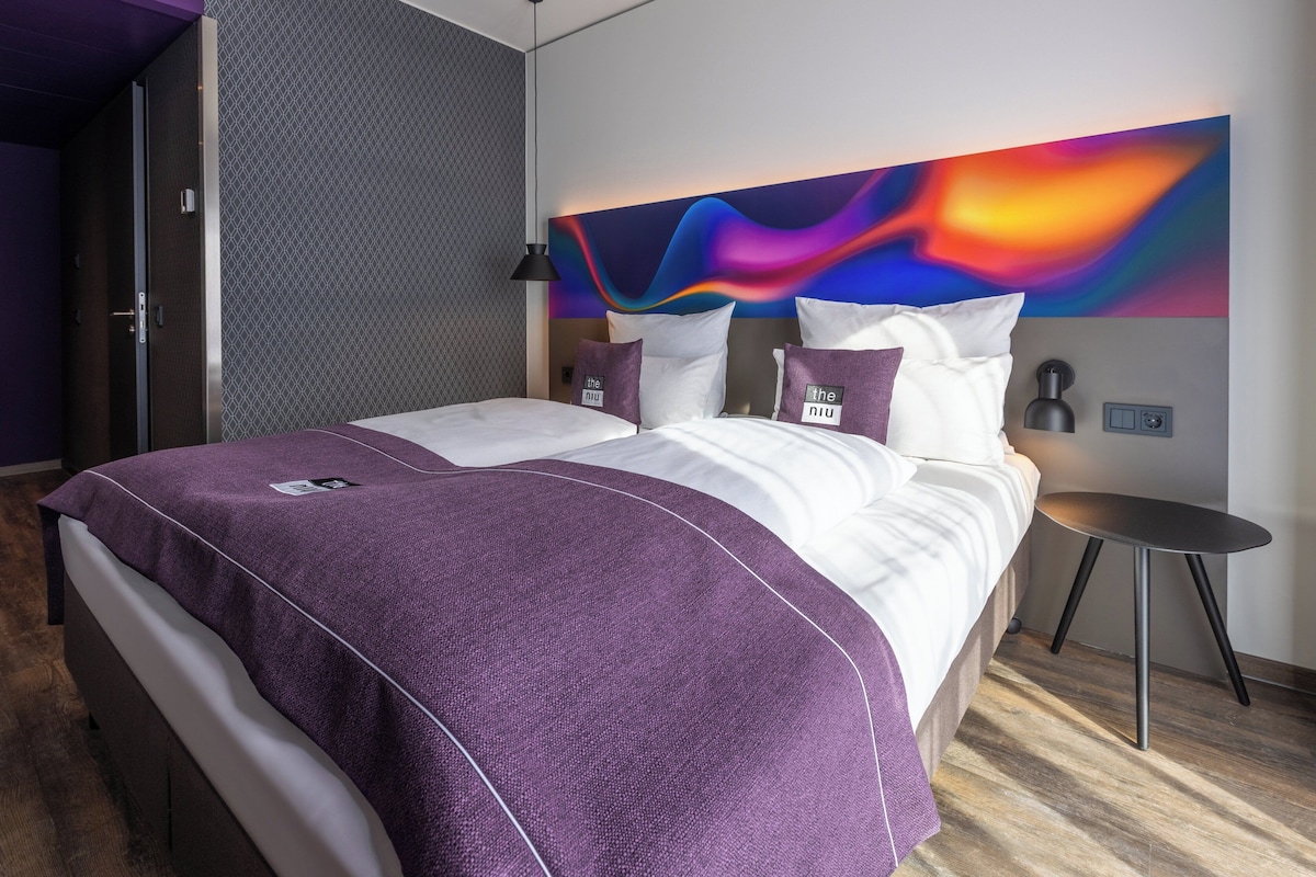 Quarto de hotel moderno com uma cama grande, uma obra de arte abstrata vibrante e uma pequena mesa de apoio.