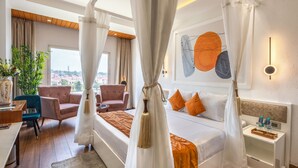 Quarto de hotel com uma cama grande, área de estar com uma poltrona e uma mesa, mesa de cabeceira com abajur, e vista da cidade pela janela.