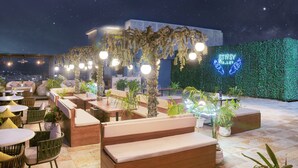 Uma área externa com bancos e mesas estofados, rodeada por plantas em vasos e um letreiro neon.