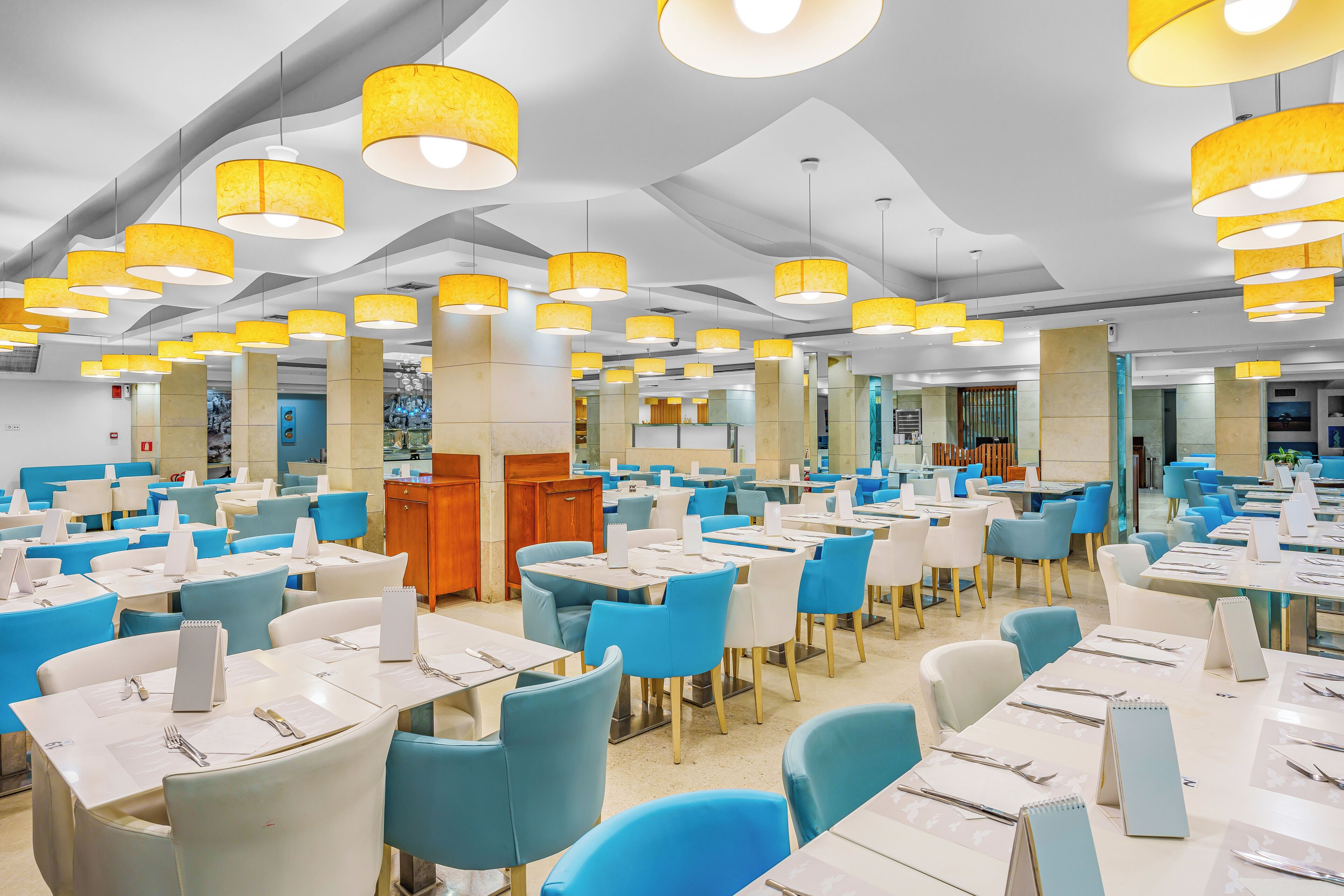 Uma ampla sala de jantar com várias mesas e cadeiras, luminárias pendentes amarelas e um balcão central.