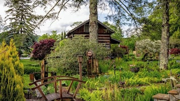 Uma cabana rústica rodeada por vegetação exuberante, uma ponte de madeira, um jardim com diversas plantas e um pequeno lago.