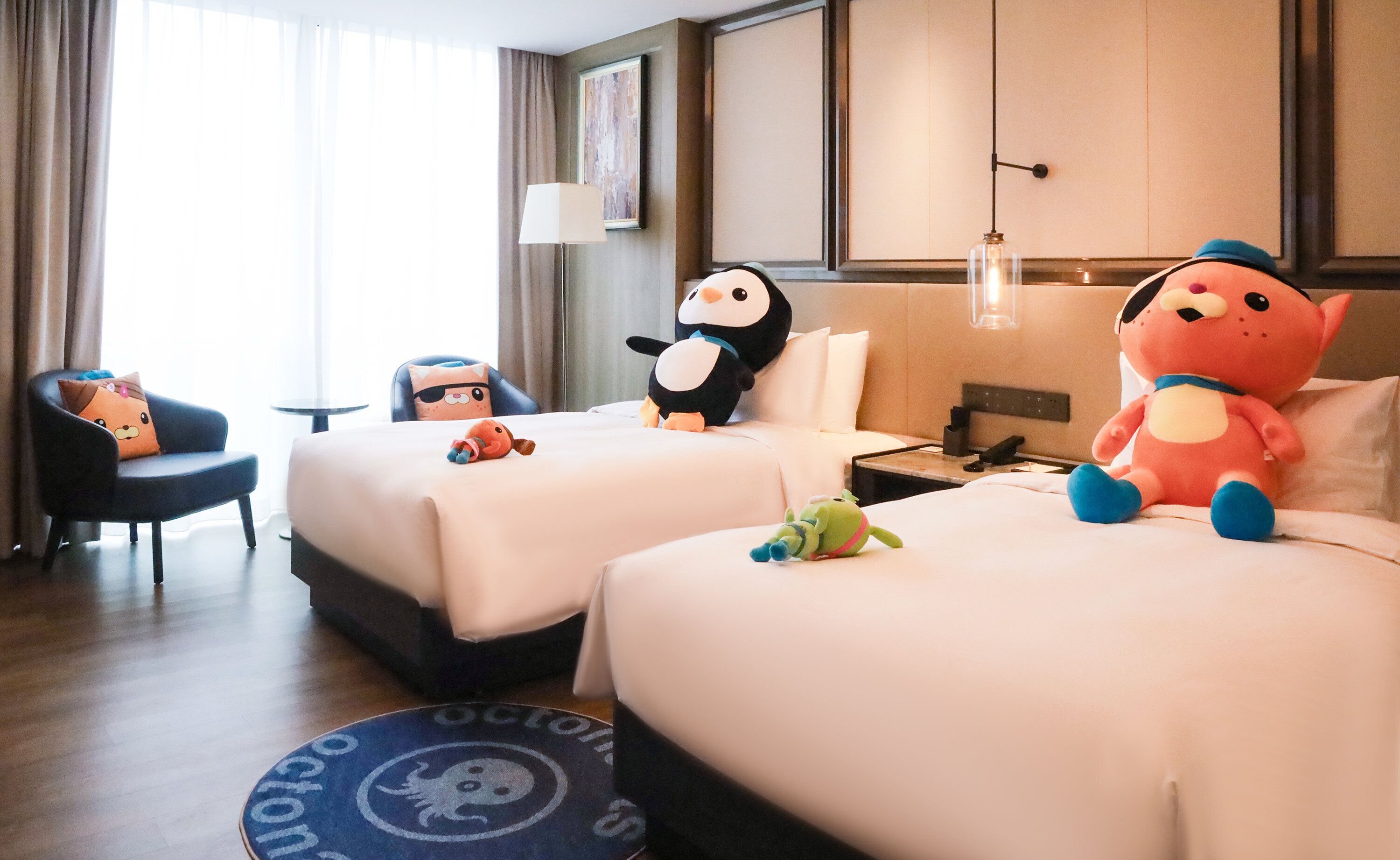 Quarto de hotel com duas camas, uma cadeira, uma mesa pequena e um abajur.
