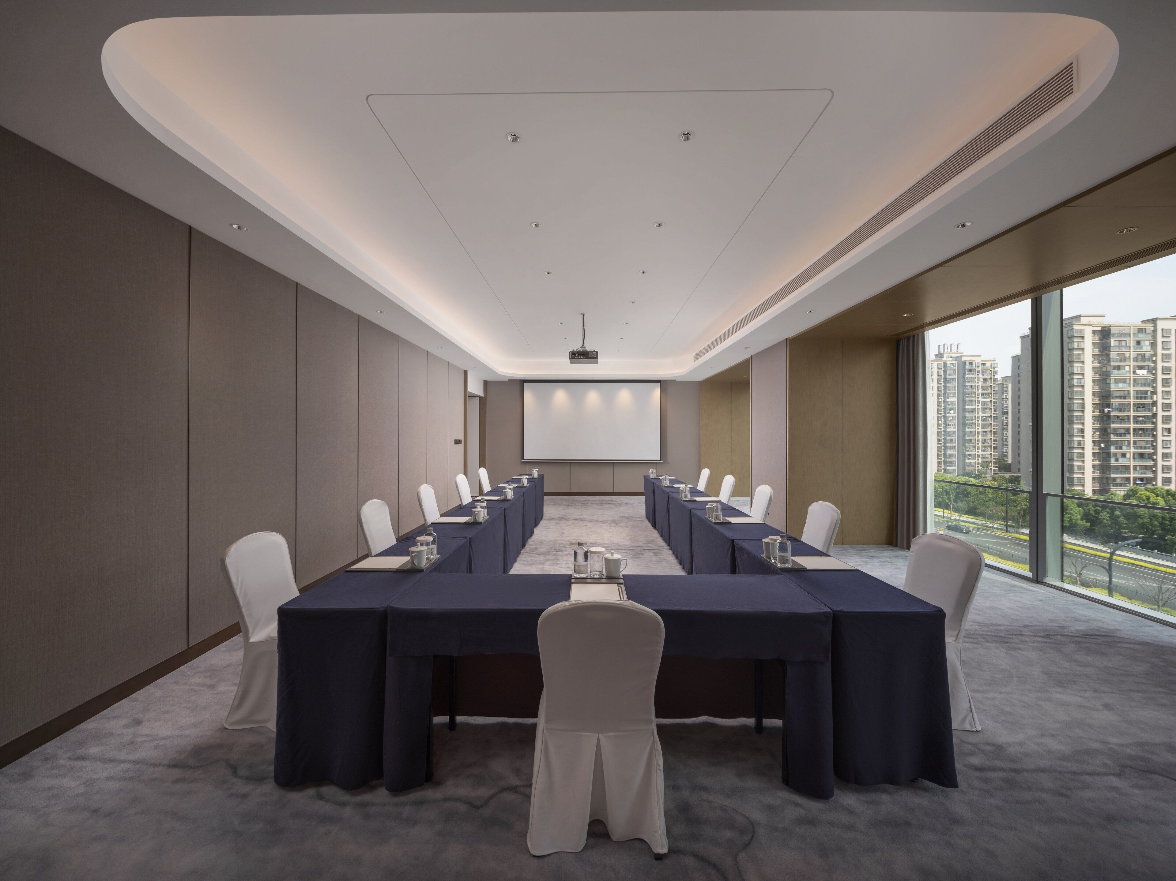 Uma sala de conferências com uma mesa comprida, cadeiras brancas, tela de projetor e janelas amplas com vista para a cidade.