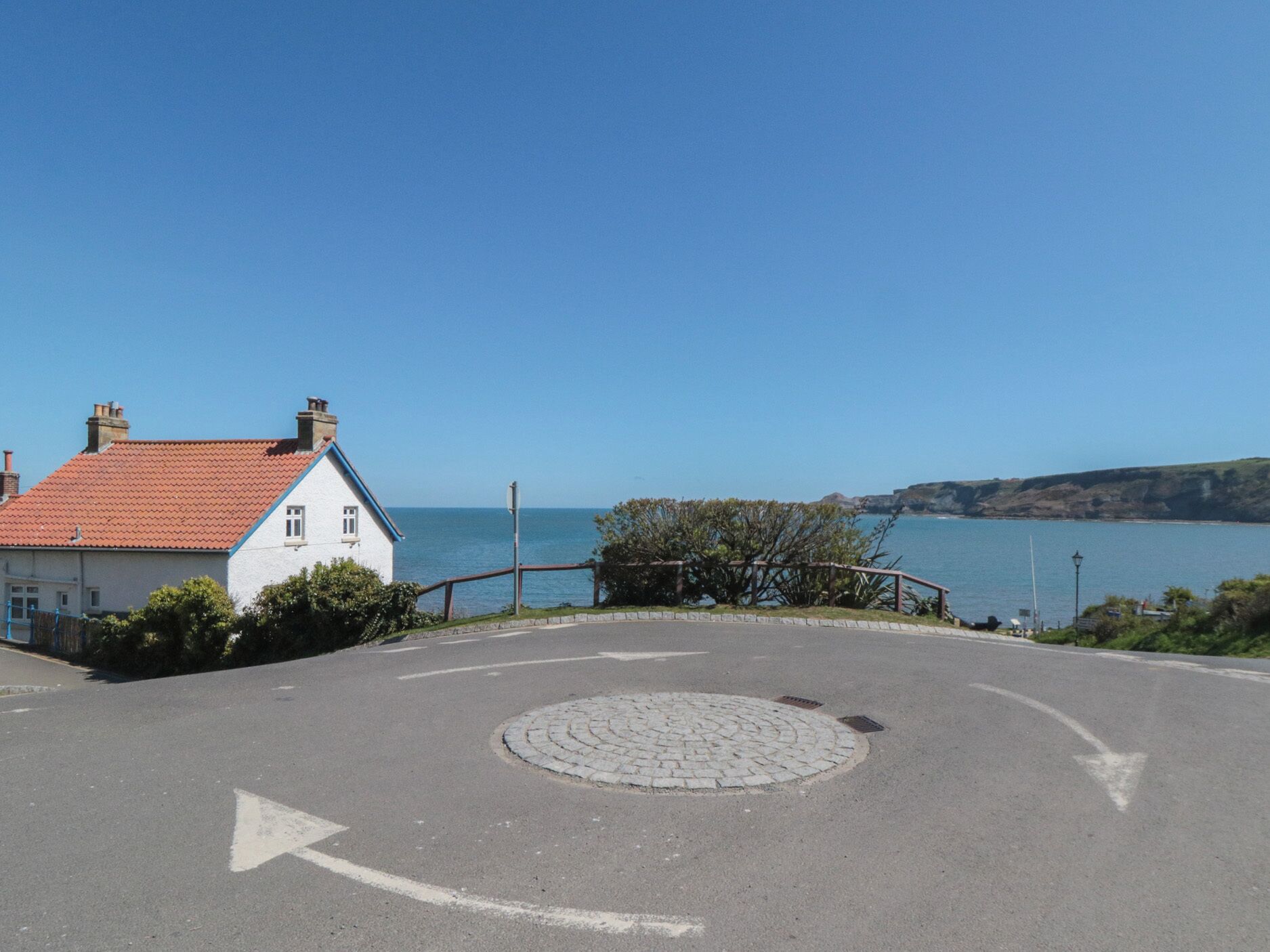 Uma estrada costeira com uma rotatória, um edifício branco com telhado vermelho e vista para o mar.