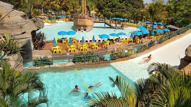 Uma grande piscina ao ar livre com um escorregador em formato de navio, cercada por palmeiras e cadeiras de sol amarelas.
