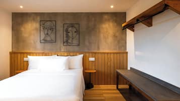 Quarto com cabeceira de madeira, cama com roupa de cama branca, um banquinho e duas obras de arte penduradas na parede.