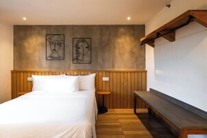 Quarto com cabeceira de madeira, cama com roupa de cama branca, um banquinho e duas obras de arte penduradas na parede.