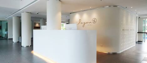 Uma área de recepção de hotel moderna, com um balcão de atendimento branco e uma parede curva exibindo o nome do hotel.