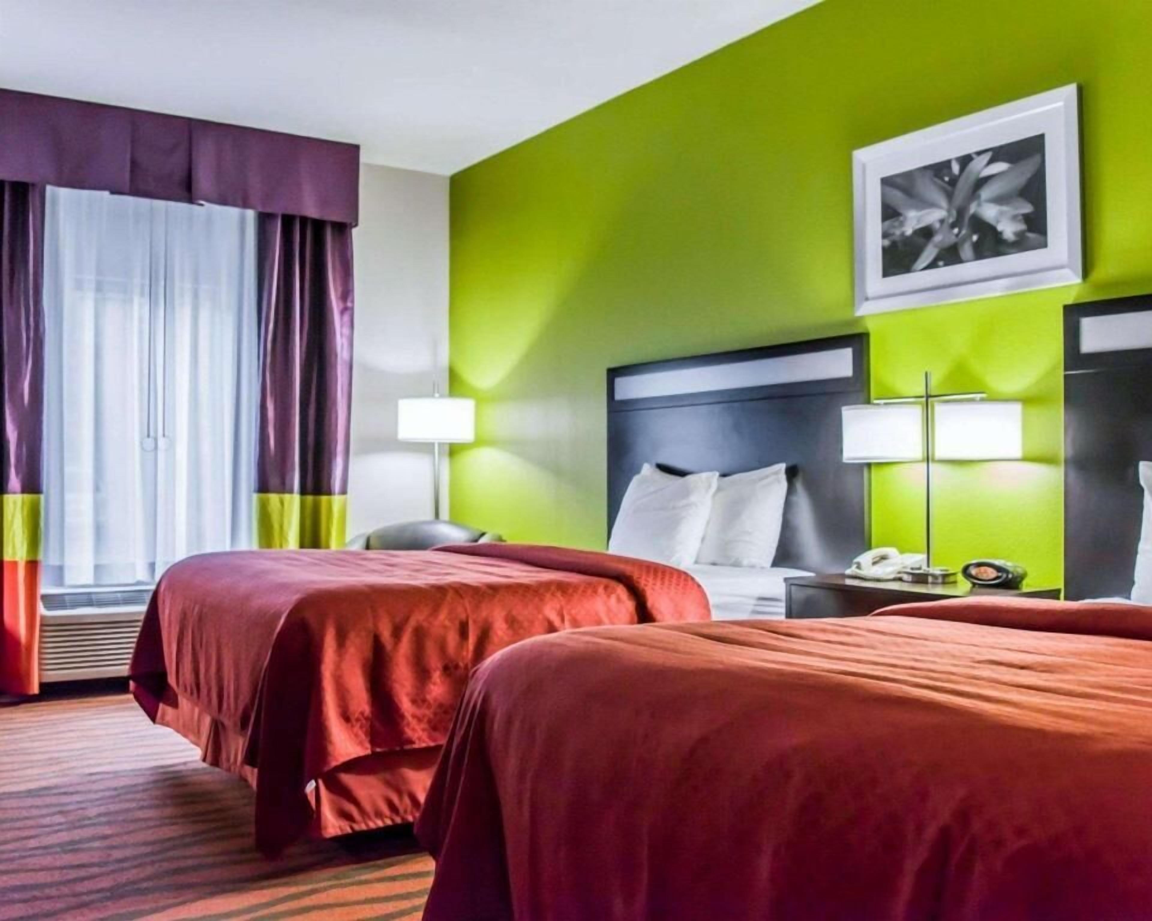 Quarto de hotel com duas camas, uma janela grande com cortinas, uma parede de destaque verde e um quadro emoldurado.