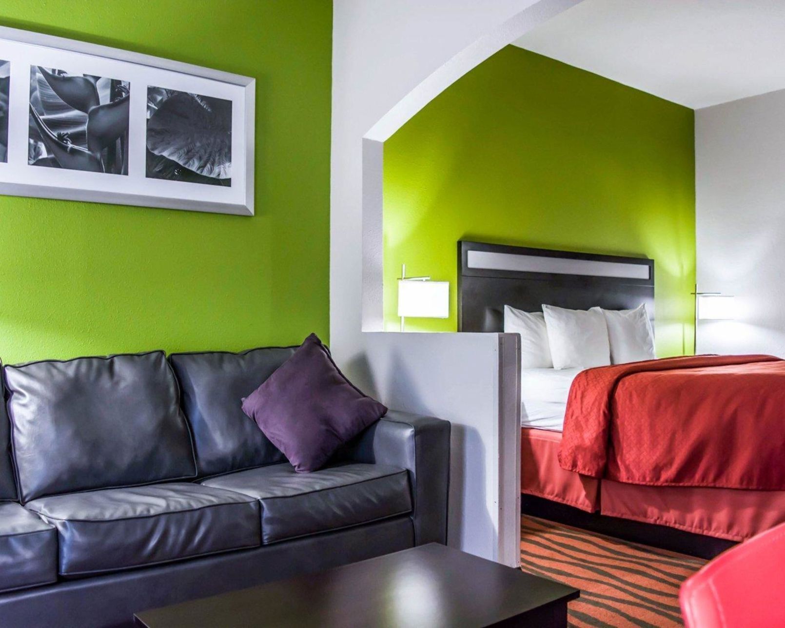 Quarto de hotel com parede verde, sofá de couro preto, cama com roupa de cama vermelha e mesa de cabeceira.
